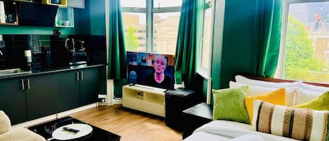 Quarto de hotel moderno com cozinha compacta, televisão, cama com várias almofadas e vista da cidade através de grandes janelas.