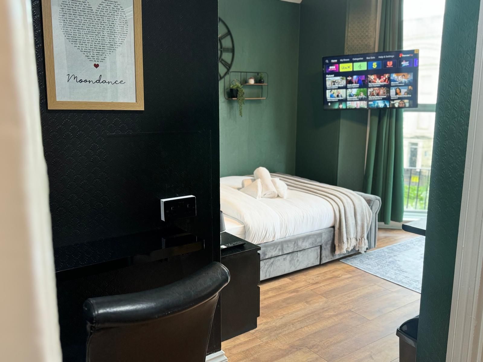 Quarto de hotel com cama, escrivaninha, cadeira, televisão presa à parede e janela com cortinas.