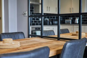 Uma sala de jantar com mesa de madeira e cadeiras pretas, um conjunto de sousbicos e vista para uma cozinha através de uma divisória de vidro.