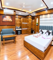 Quarto de hotel com piso de madeira, cama com colcha branca, sofá, escrivaninha e ventilador de teto.