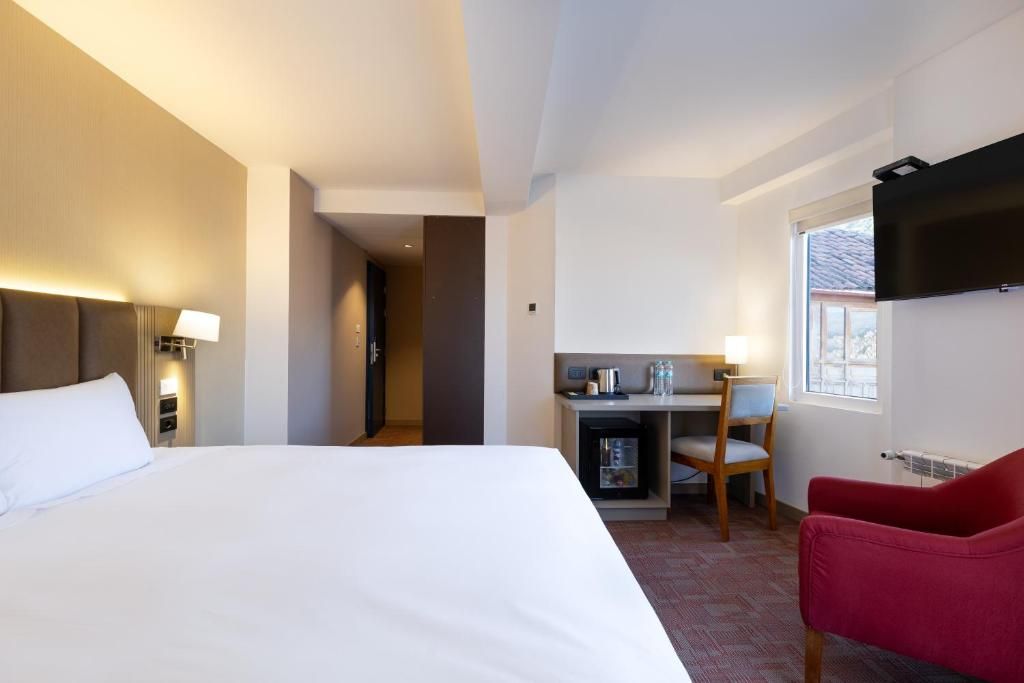 Quarto de hotel com cama, escrivaninha, cadeira e televisão.