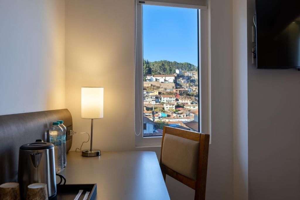 Quarto de hotel com vista para uma cidade, uma mesa com abajur, uma cadeira e uma TV na parede.