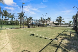 Uma quadra de tênis com rede, cercada por uma grade, palmeiras e edifícios ao fundo.