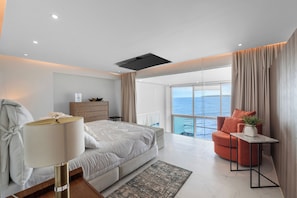 Quarto moderno com cama grande, área de estar, cômoda e vista para o mar.