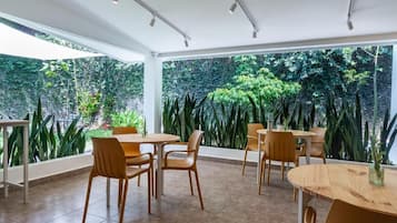 Um espaço de refeições interno com mesas e cadeiras, janelas amplas e vista para vegetação exuberante.
