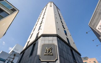 Um edifício alto com um letreiro chamativo 'MUSE' na base, cercado por outros edifícios modernos.