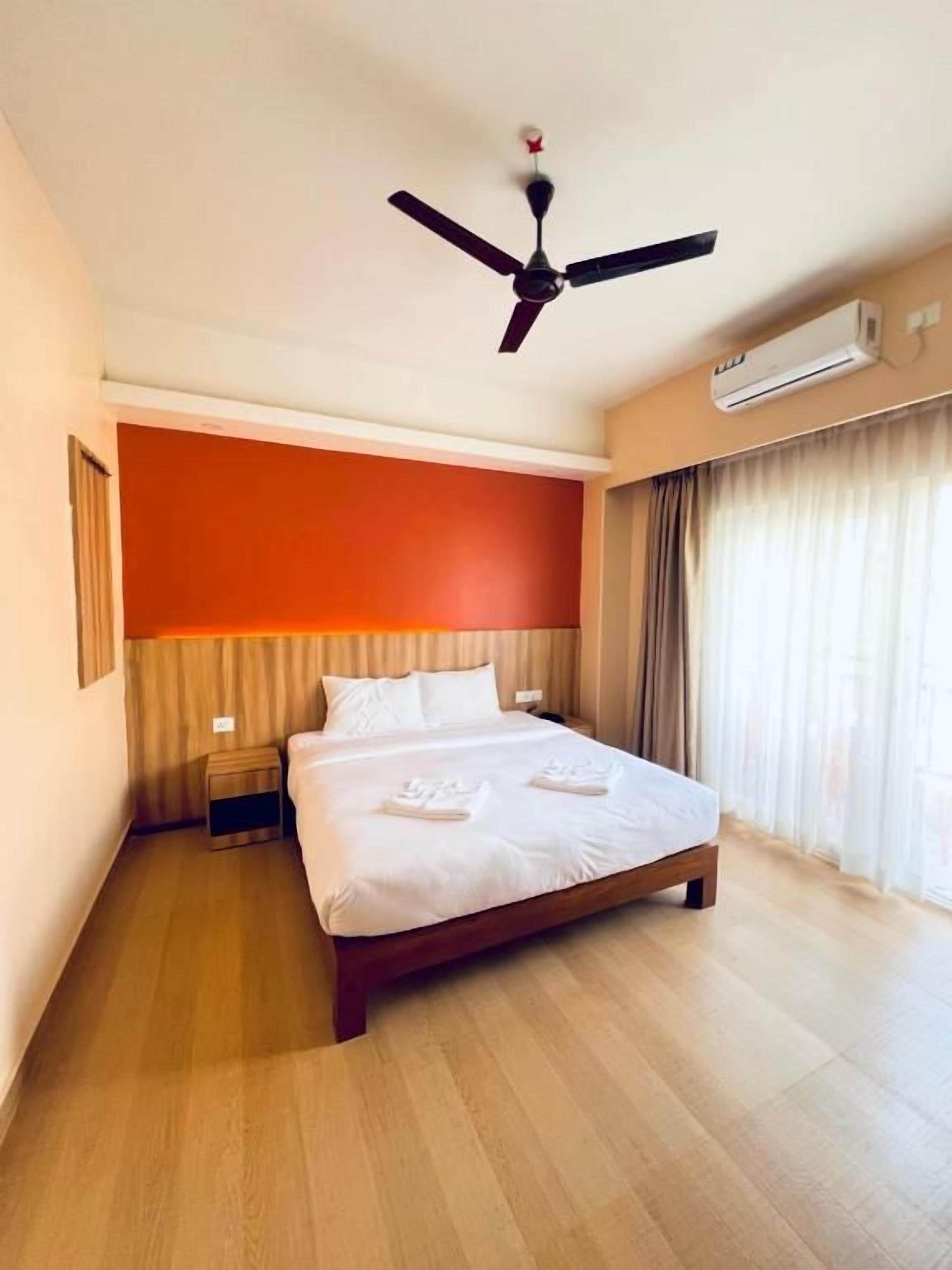 Quarto de hotel com cama, ventoinha no teto, ar condicionado e janela com cortinas.