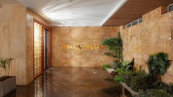 Um hall com piso lustroso, parede de pedra com inscrição dourada e um pequeno jardim interno com diversas plantas.