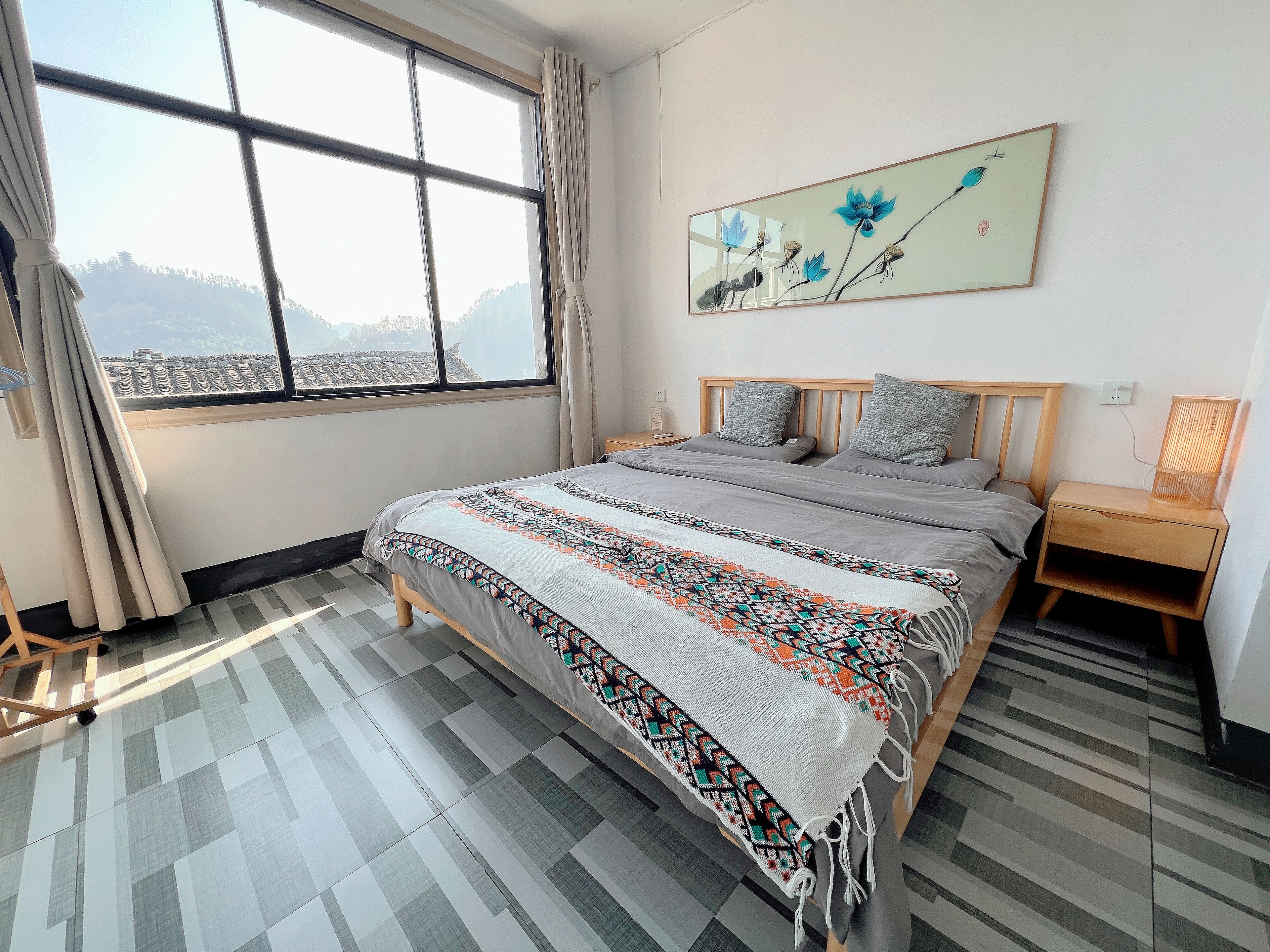 Quarto de hotel com uma janela ampla, uma cama com colcha estampada, uma mesinha de cabeceira com abajur e uma obra de arte na parede.