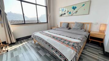 Quarto de hotel com uma janela ampla, uma cama com colcha estampada, uma mesinha de cabeceira com abajur e uma obra de arte na parede.
