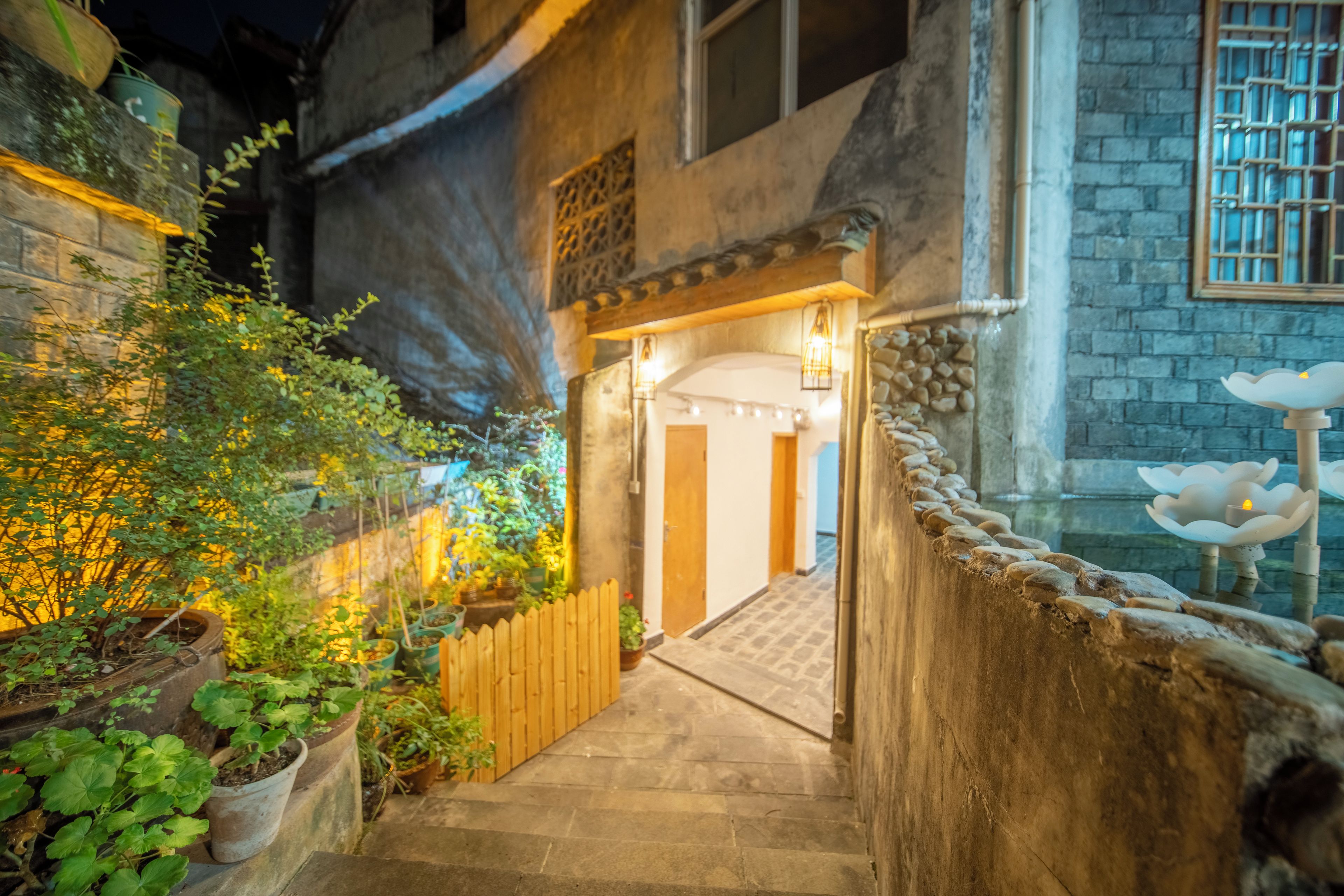 Um caminho de pedra estreito conduz a uma porta de madeira, ladeado por plantas em vasos e um muro de pedra com grade decorativa.
