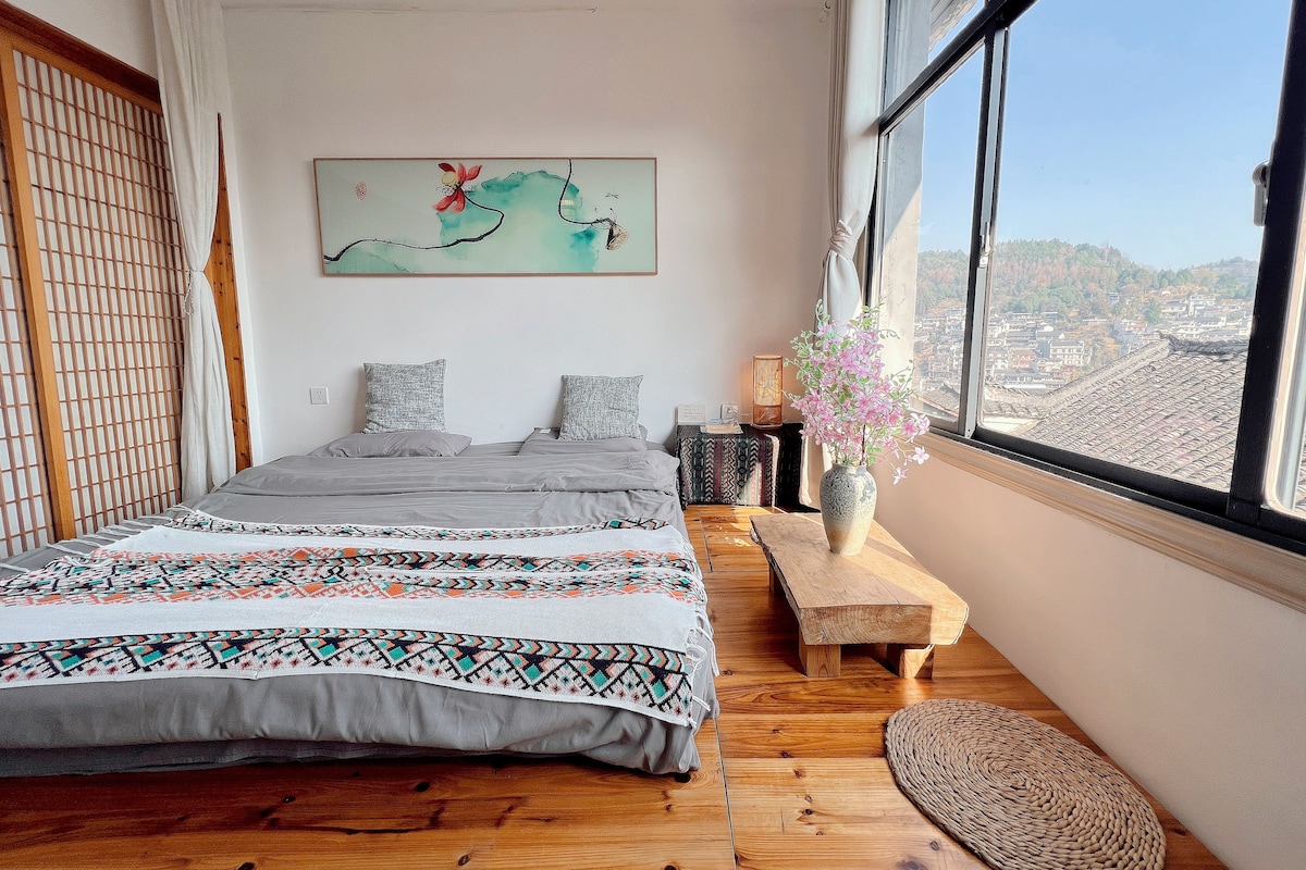 Quarto com cama, janela com vista, piso de madeira e obra de arte na parede.