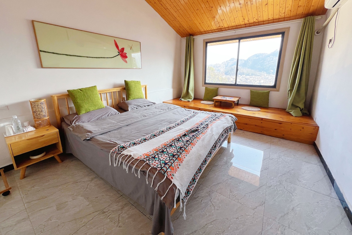Quarto com cama, cabeceira de madeira, mesas de cabeceira, janela com cortinas e vista para o exterior.