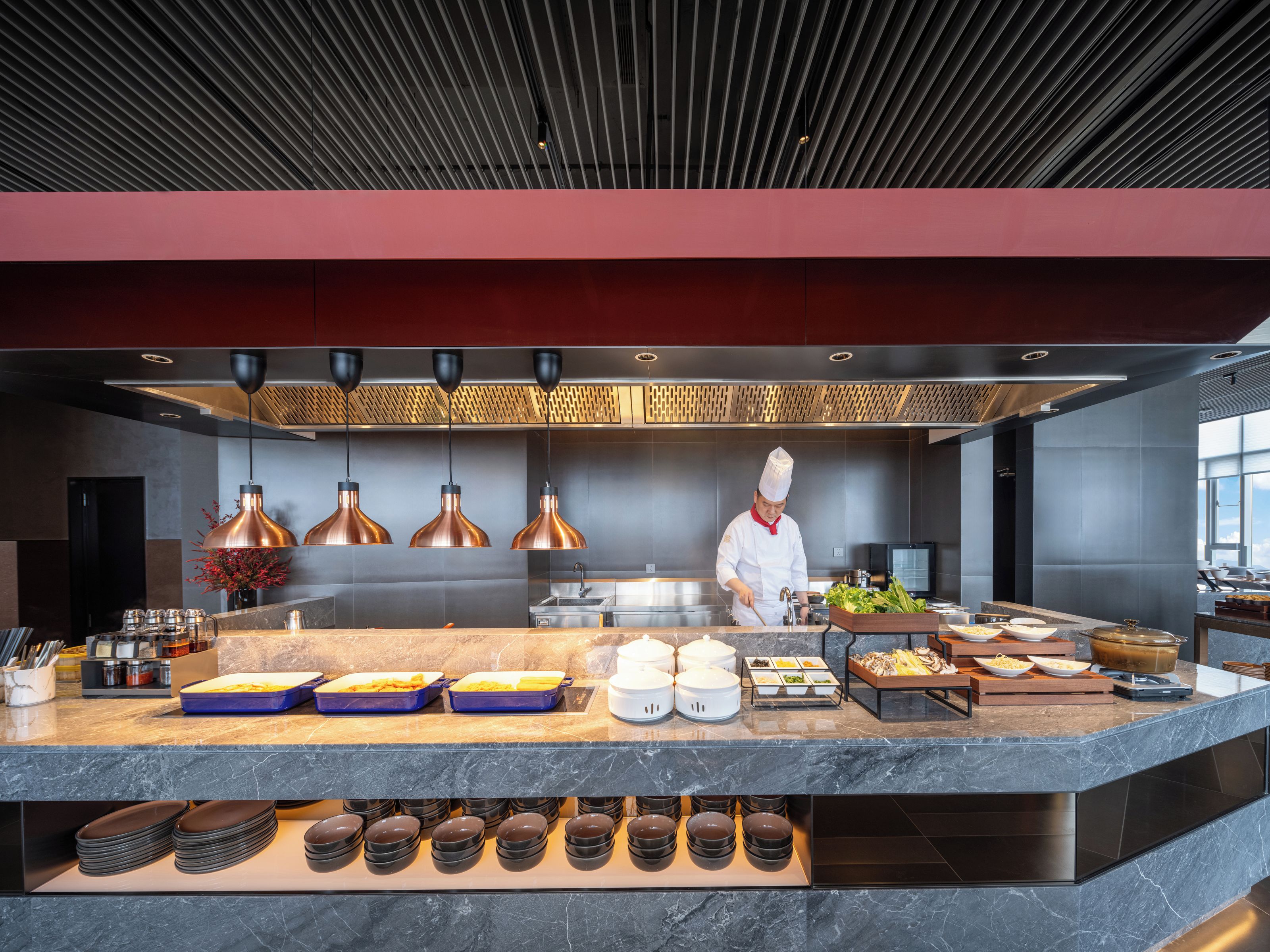 Cozinha moderna com um chef preparando comida, buffet com diversos pratos e variedade de eletrodomésticos.