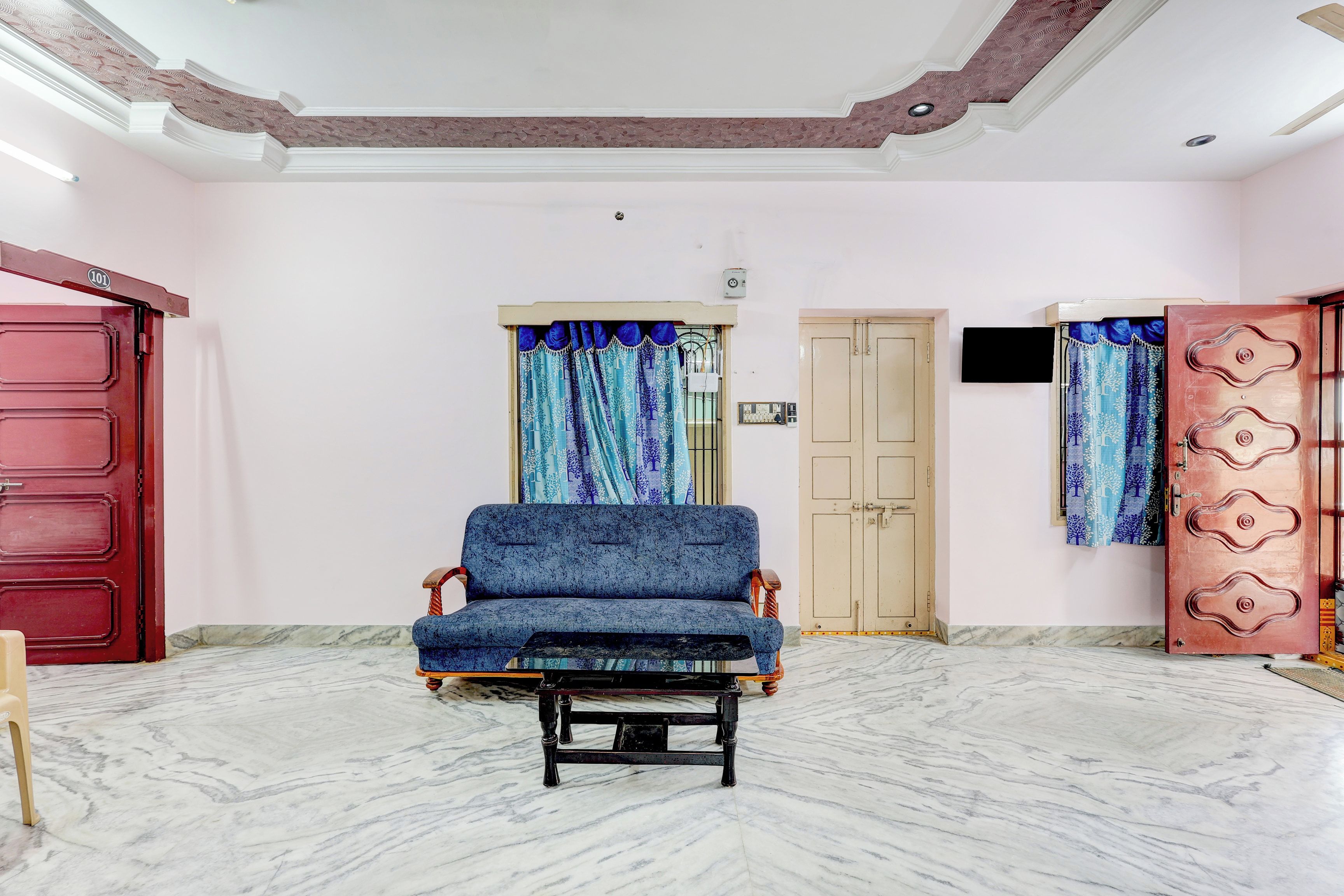 Um quarto com piso de mármore, um sofá azul, uma mesa de centro de madeira, uma porta vermelha e uma porta bege.