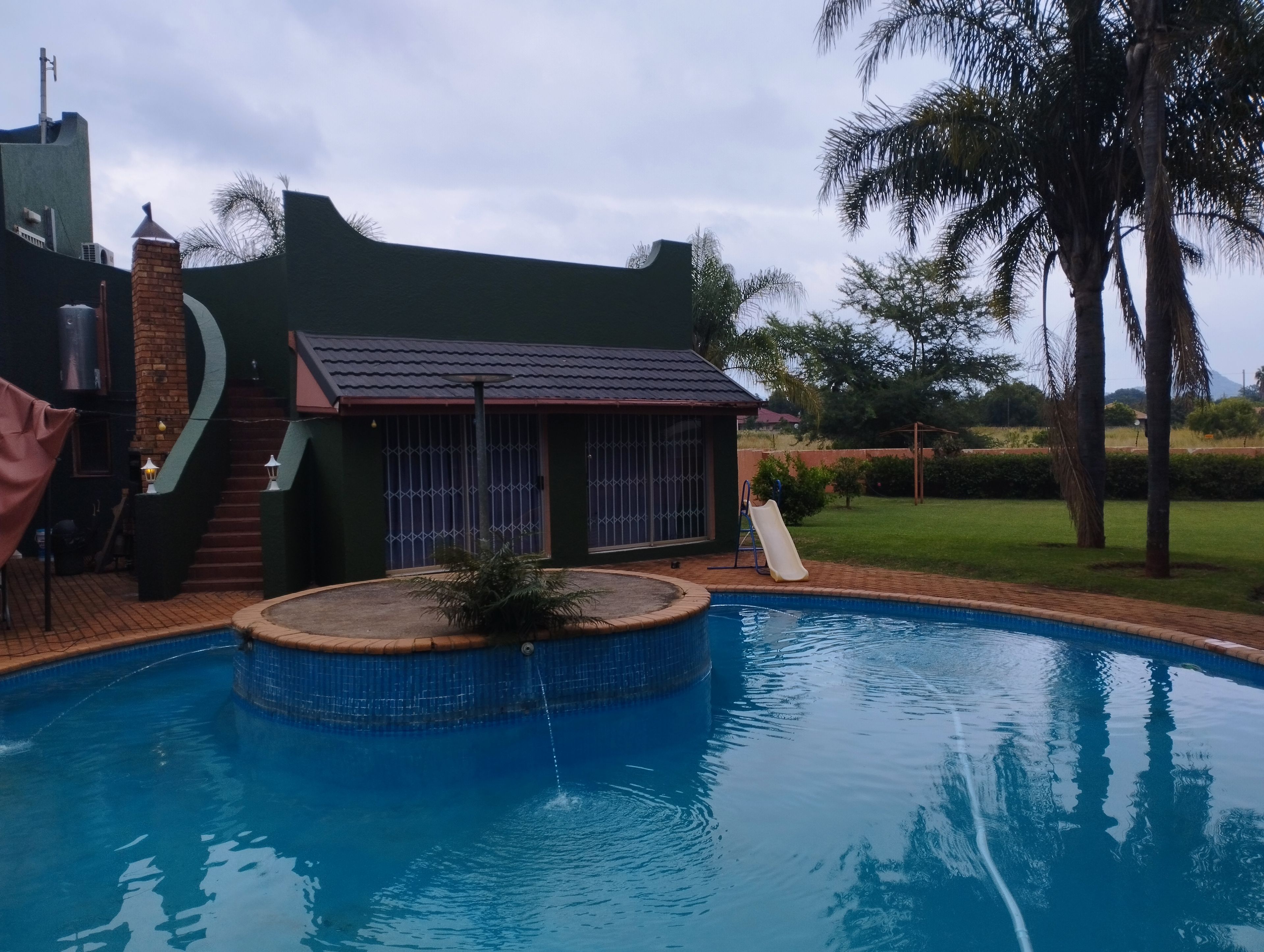 Piscina com uma pequena fonte, rodeada por um deck de tijolos e um edifício verde com telhado em azulejos.