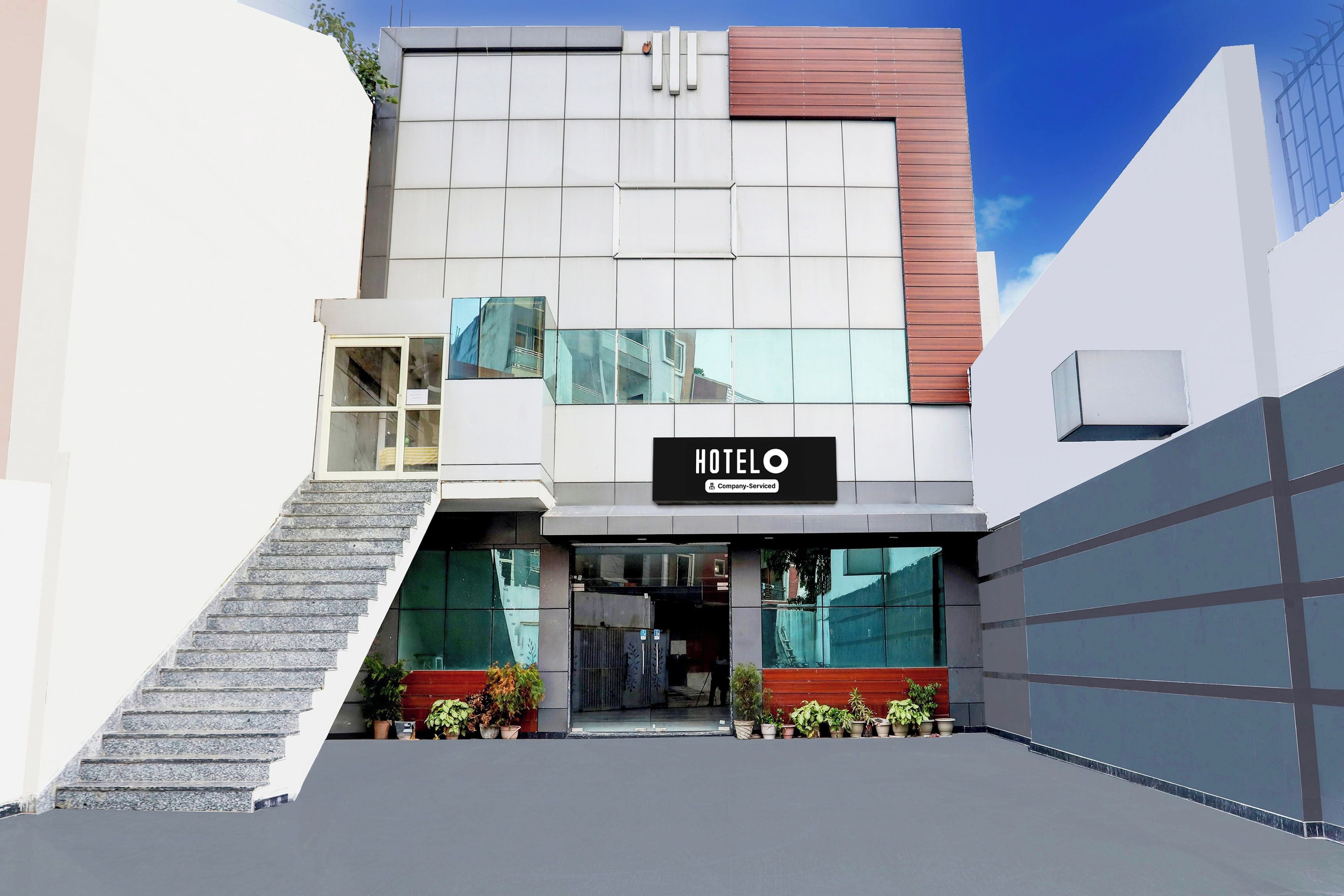 Um hotel moderno com um letreiro chamativo 'HOTEL', uma escadaria que leva à entrada e plantas em vasos na base.