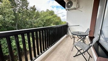 Uma varanda com guarda-corrimão preto, uma mesa com cadeiras e uma janela com vista para árvores.