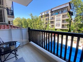 Varanda com mesa e cadeiras de vidro, com vista para uma piscina e edifícios próximos.