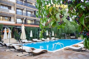 Piscina com espregui çadeiras e guarda-sóis, cercada por vegetação e um edifício com varandas.