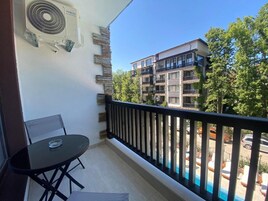 Varanda com mesa e cadeiras, piscina ao fundo e um edifício com várias varandas.