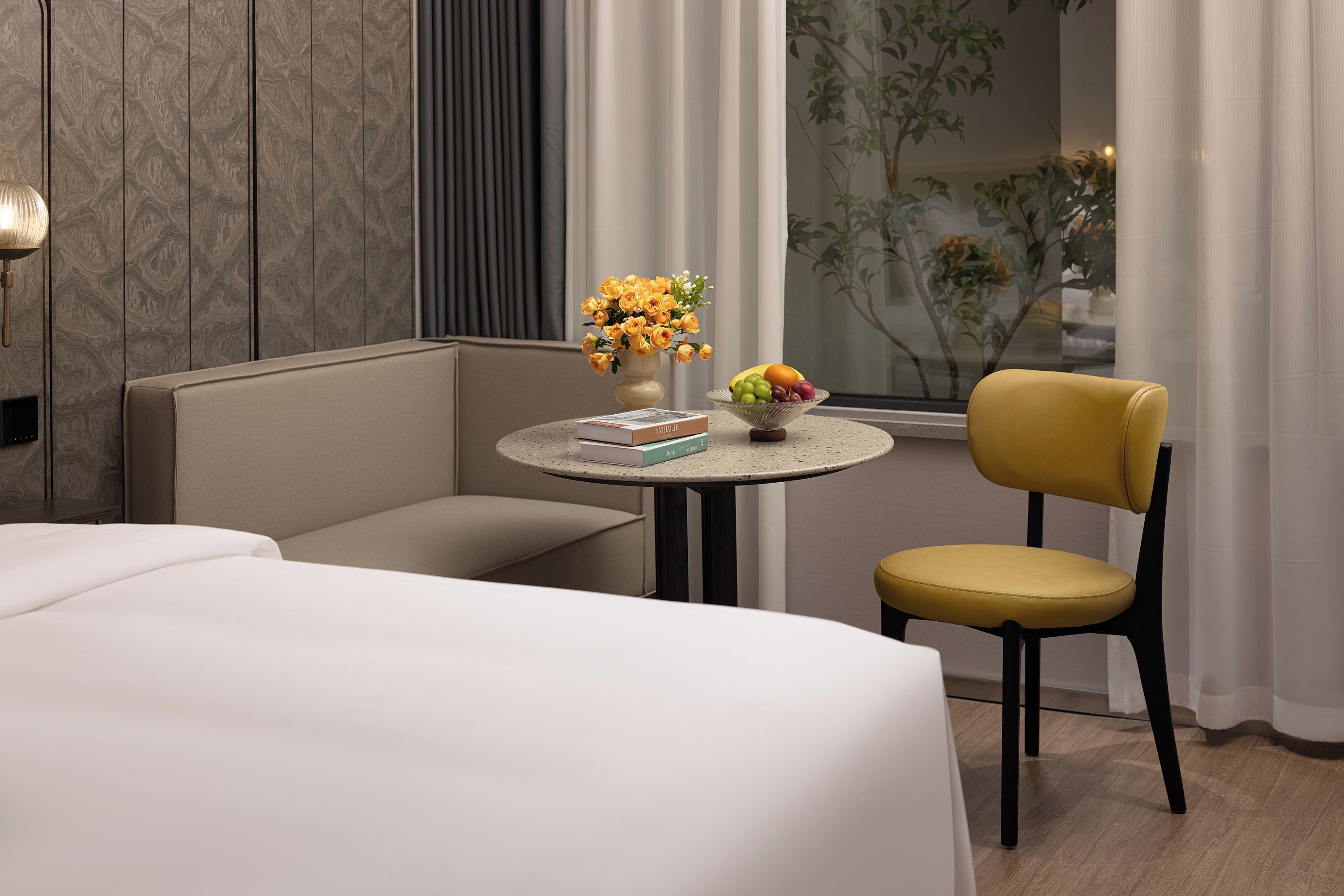 Quarto de hotel com cama, mesa redonda com vaso de flores, uma cadeira e janela com cortinas.