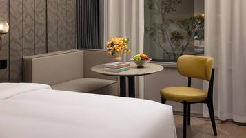 Quarto de hotel com cama, mesa redonda com vaso de flores, uma cadeira e janela com cortinas.