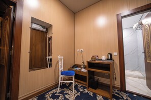 Quarto de hotel com porta de madeira, uma escrivaninha com cadeira, uma mesa pequena com chaleira, lixeira e espelho.