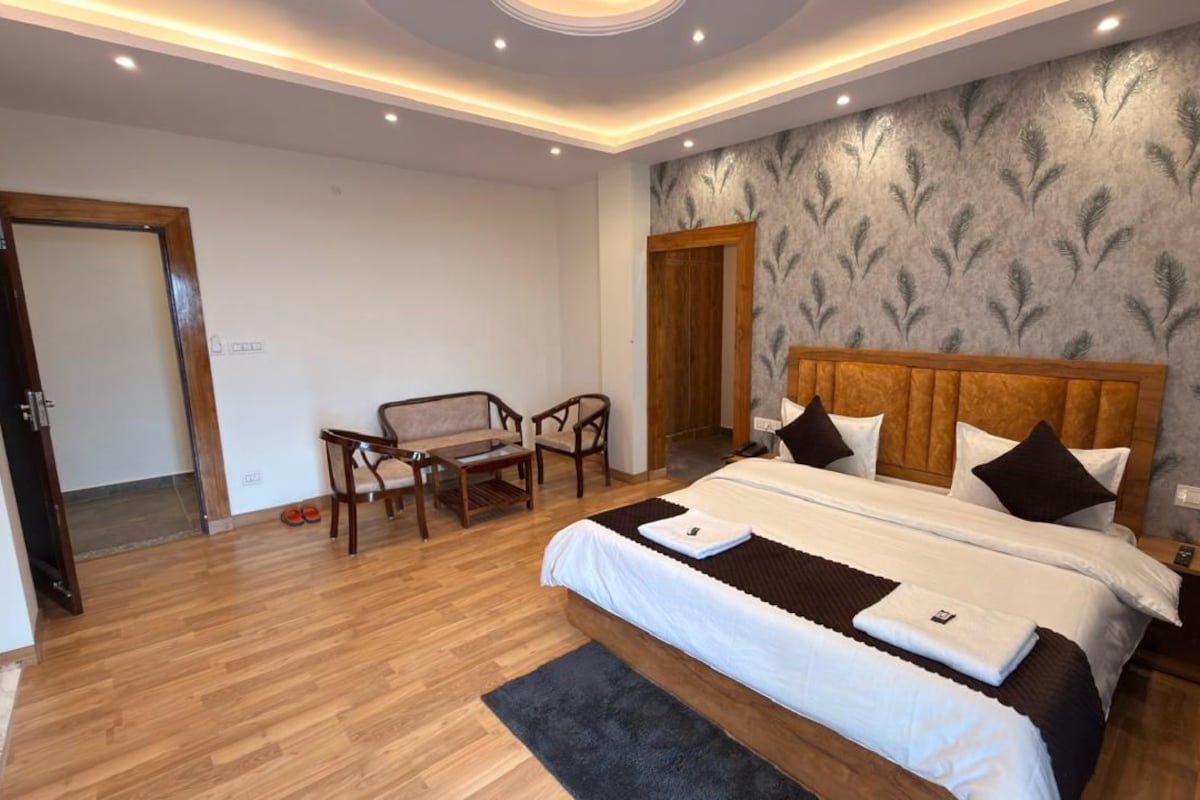 Quarto de hotel com uma cama grande, cabeceira de madeira, área de estar com cadeiras e parede estampada.