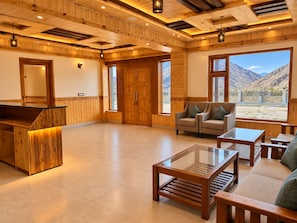 Quarto amplo com painéis de madeira, balcão de recepção, área de estar com sofás e mesas, e grandes janelas com vista para as montanhas.