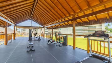 Uma área de ginástica ao ar livre com equipamentos de exercício, incluindo esteira e aparelhos de musculação, sob uma estrutura de telhado de madeira.