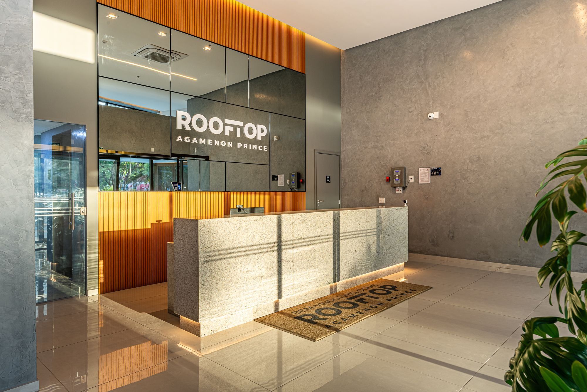 Uma mesa de recepção com divisória de vidro e uma placa com a inscrição 'ROOFTOP AGAMENON PRINCIPAL'.