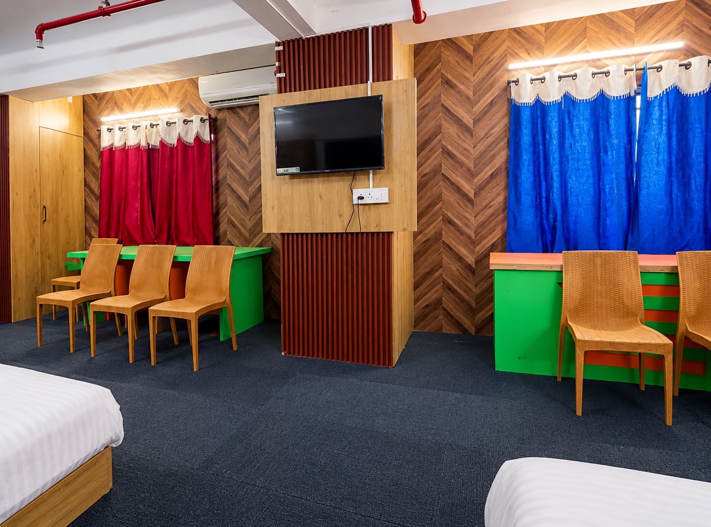 Quarto de hotel com cama, escrivaninha com cadeira, televisão na parede e janela com cortinas.