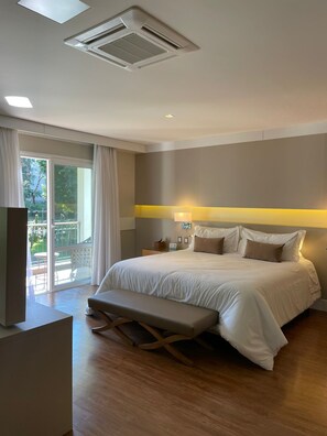 Quarto de hotel com uma cama grande, um banco, uma escrivaninha e vista pela porta de vidro.