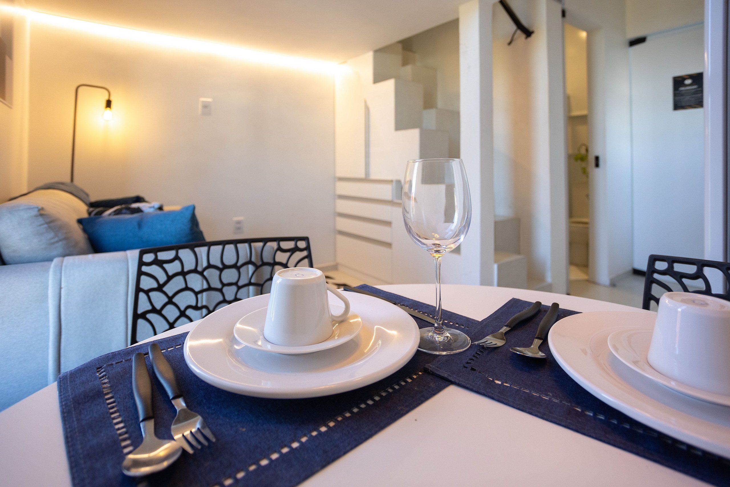 Uma área de refeições com uma mesa posta para dois, incluindo pratos, uma xícara, uma taça de vinho e talheres.