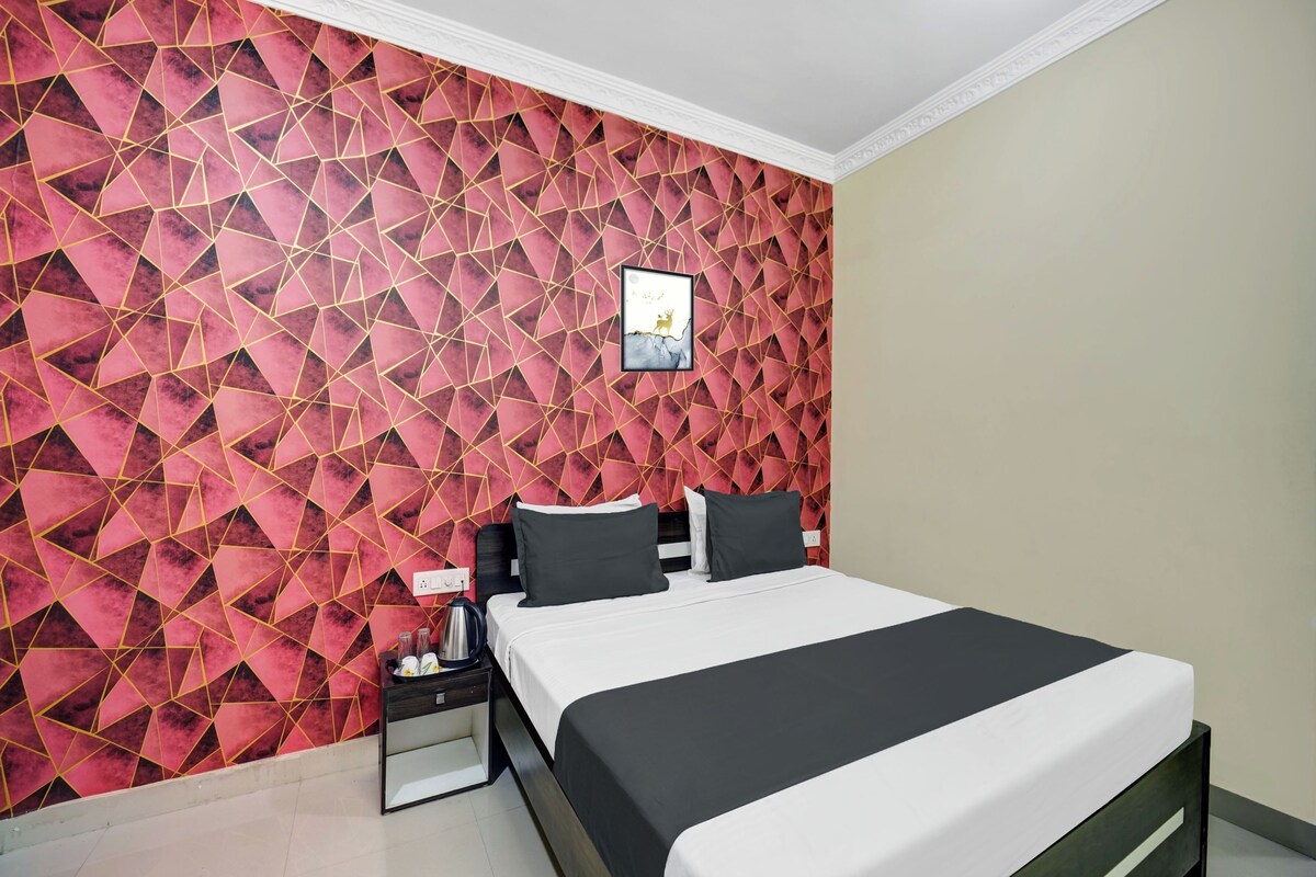 Quarto de hotel com parede estampada em padrão geométrico, cama com roupa de cama preta e branca, mesa de cabeceira com chaleira e quadro pendurado na parede.