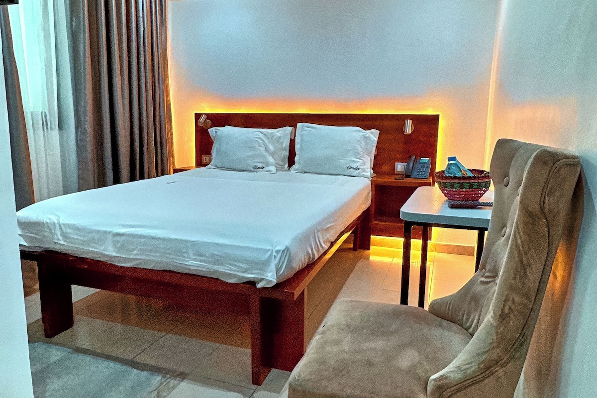 Quarto de hotel com uma cama, uma cadeira, uma mesa, aparelho de ar condicionado e mesa de cabeceira.