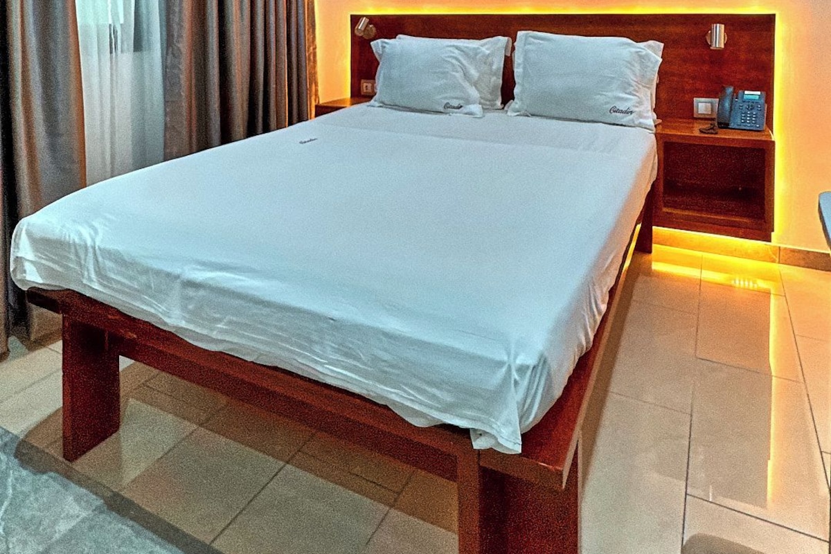 Quarto de hotel com uma cama grande, ar condicionado, mesa de cabeceira com telefone e criado-mudo.