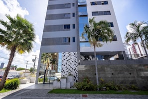Um edifício moderno de vários andares com fachada geométrica, entrada de vidro e áreas verdes ao redor.