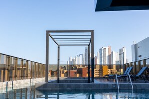 Uma piscina no terraço com guarda-corpo de vidro, espreguiçadeiras e um horizonte urbano ao fundo.