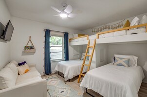 Quarto com beliches, ventoinha no teto, janela com cortinas, um pequeno sofá e televisão fixada na parede.