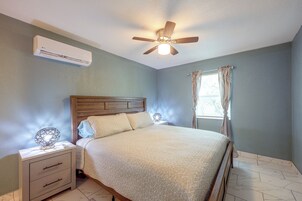 Quarto com cama, mesa de cabeceira, ventilador de teto, aparelho de ar condicionado e janela com cortinas.