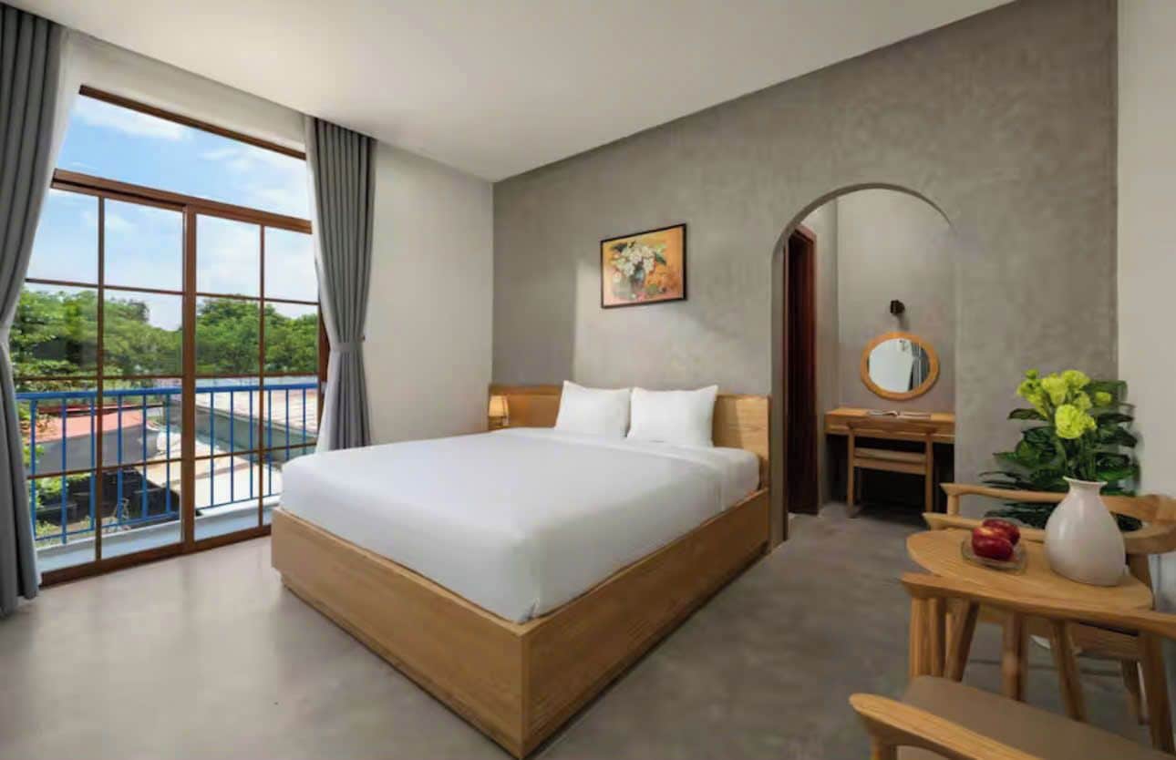 Quarto de hotel com uma cama grande, uma escrivaninha, uma cadeira, uma mesinha, um espelho e vista para o verde lá fora.