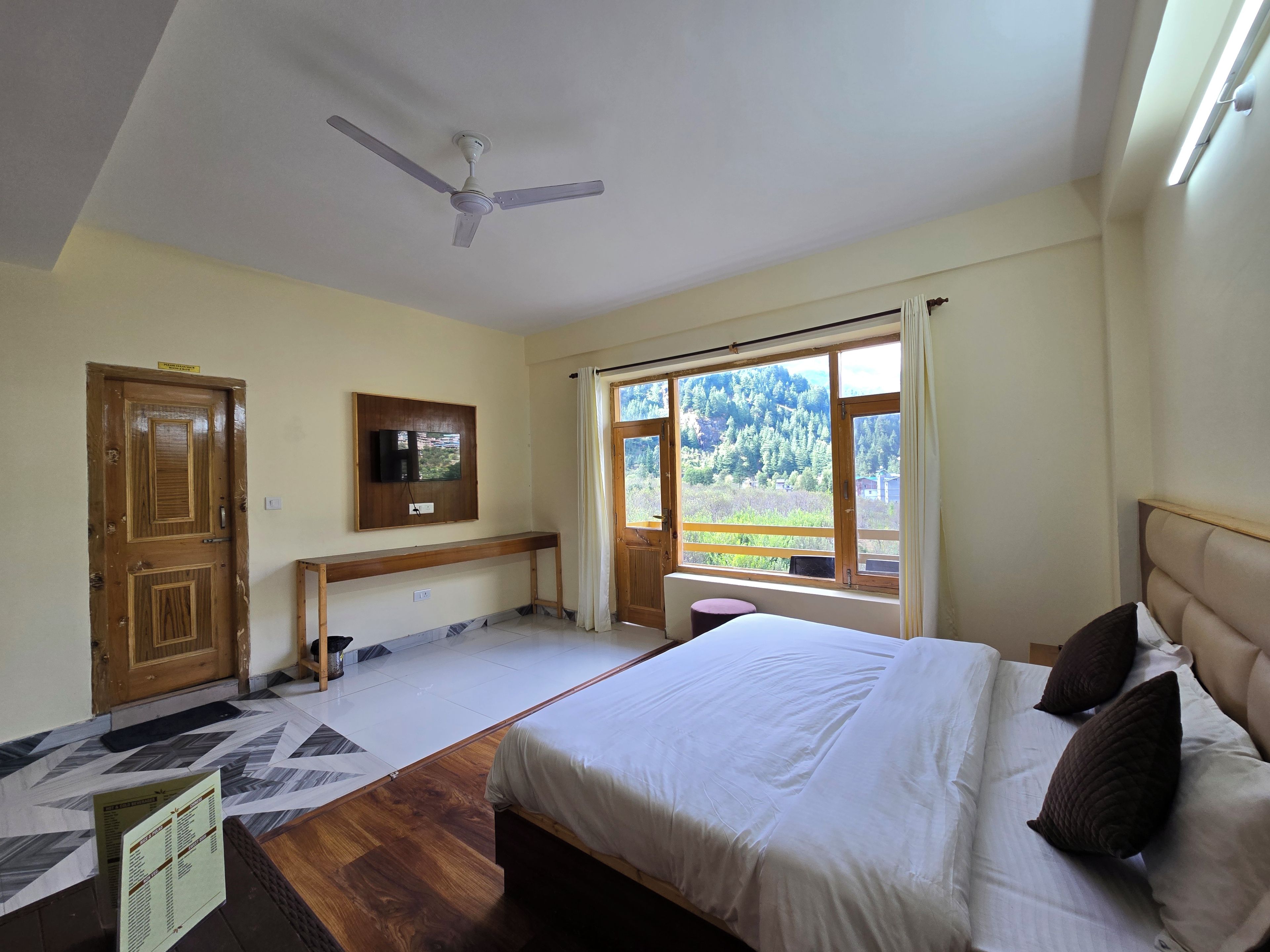 Quarto com cama, ventilador de teto, televisão na parede, janela com vista para árvores e porta de madeira.