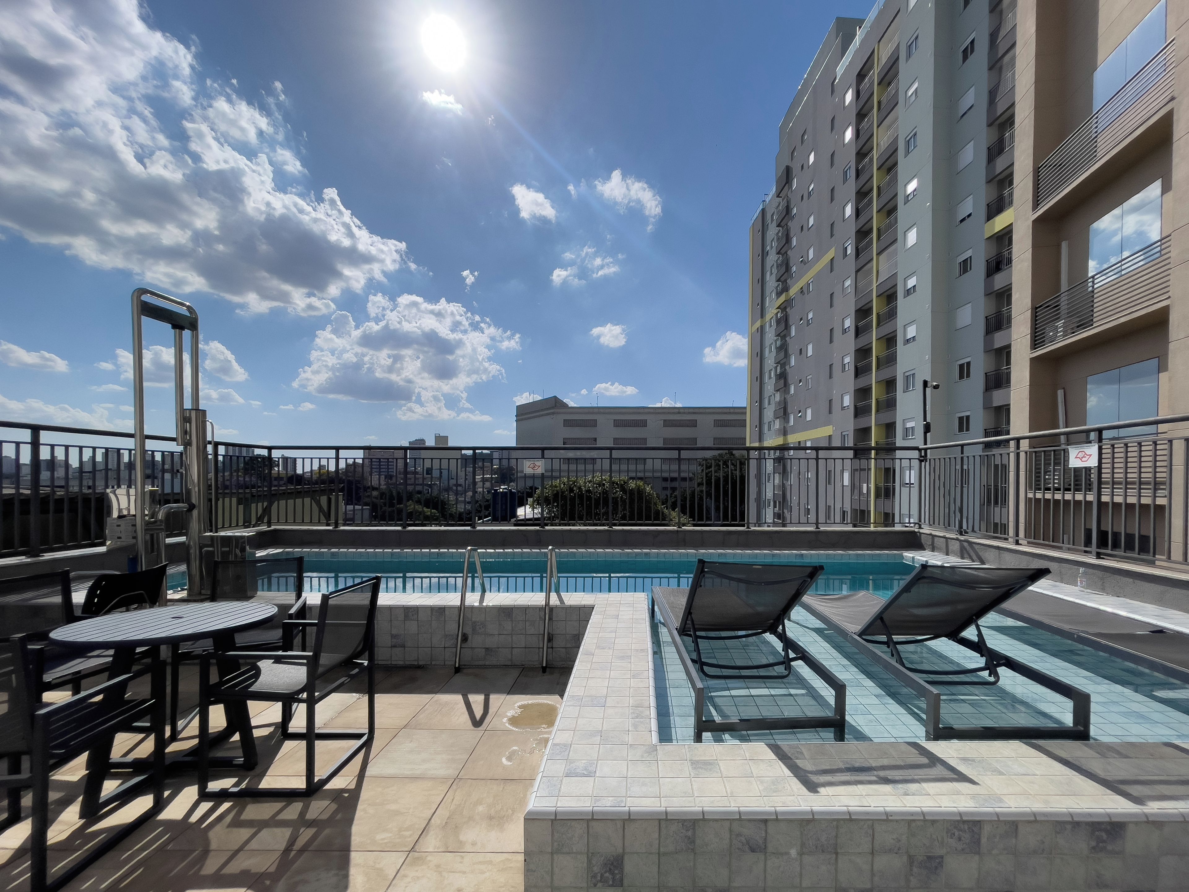 Área de piscina no terraço, com espreguiçadeiras, uma mesa e vista para o horizonte urbano.