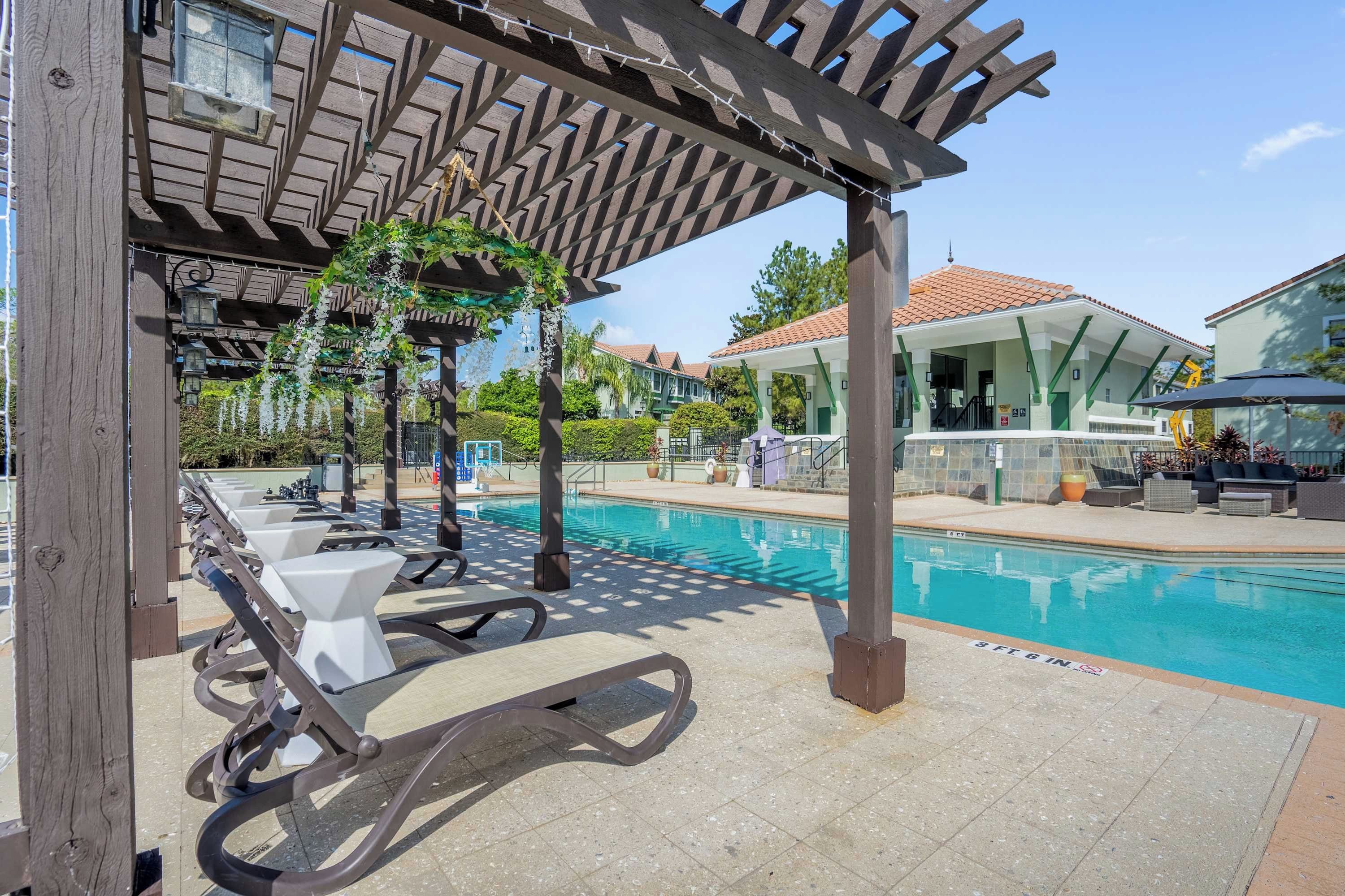 Área de piscina com espreguiçadeiras, um pérgola com vegetação e um edifício com telhado de azulejos.