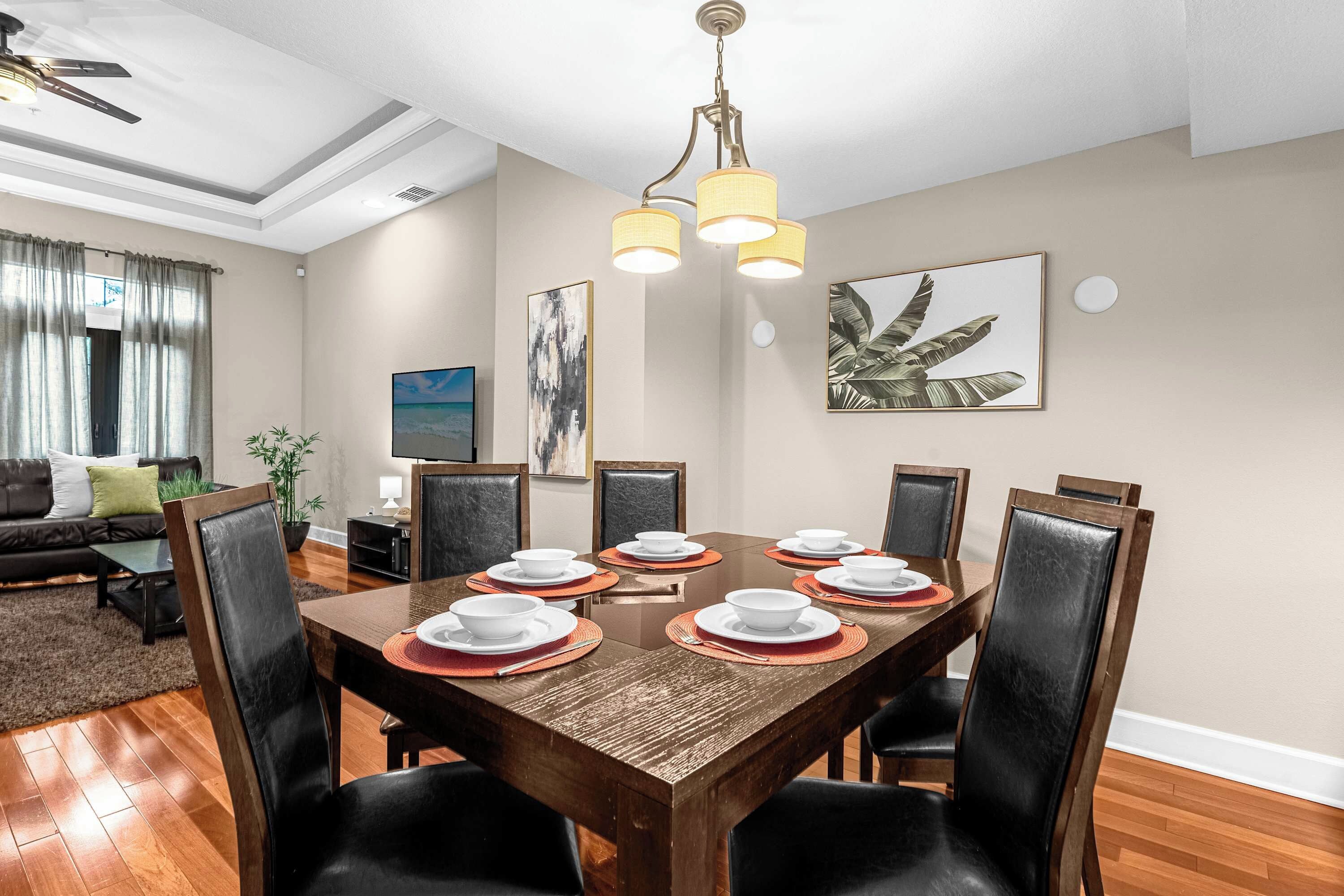 Uma área de refeições com mesa de madeira posta para seis lugares, rodeada por cadeiras pretas, com ventoinha no teto e luminárias pendentes.