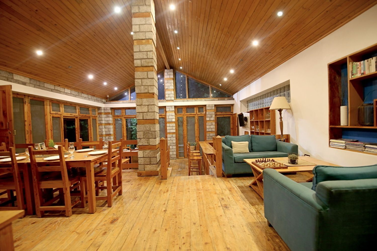 Um quarto amplo com piso de madeira, um pilar de pedra, um sofá verde, uma mesa de xadrez e uma estante de livros.