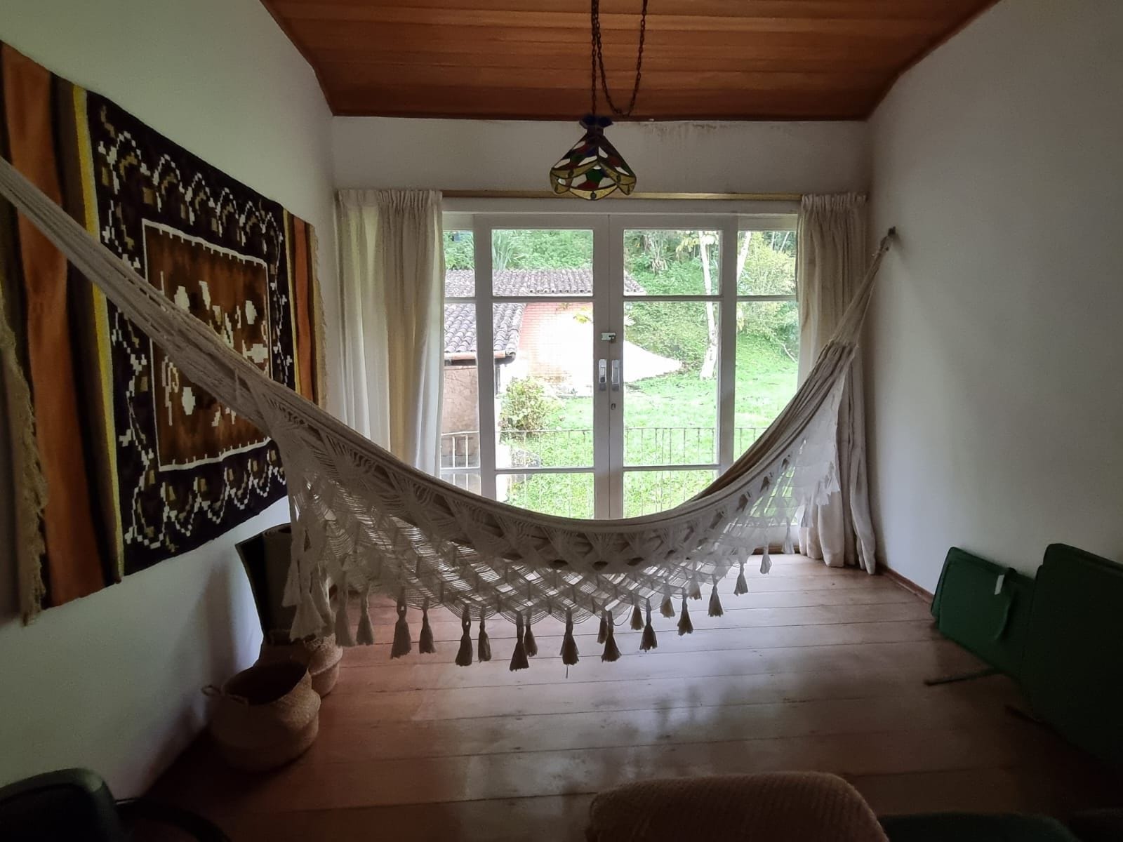 Uma rede em um quarto com teto de madeira, uma janela grande com cortinas e vista para o verde lá fora.