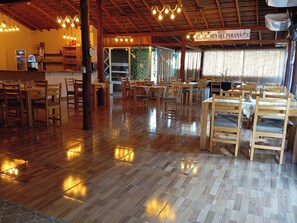 Restaurante com mesas e cadeiras de madeira, chão de azulejos e luminárias pendentes.